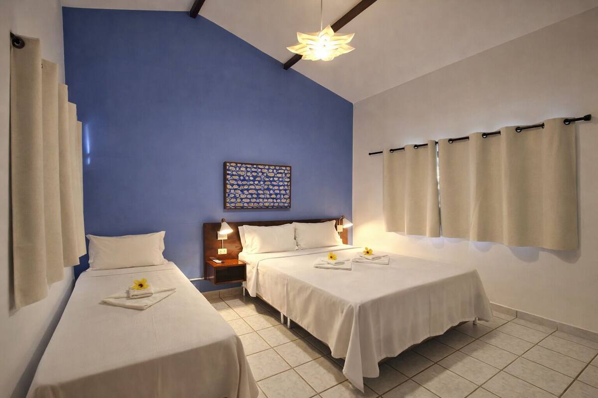 Quarto de hotel com duas camas, uma mesa de cabeceira, uma luminária na parede e iluminação no teto.
