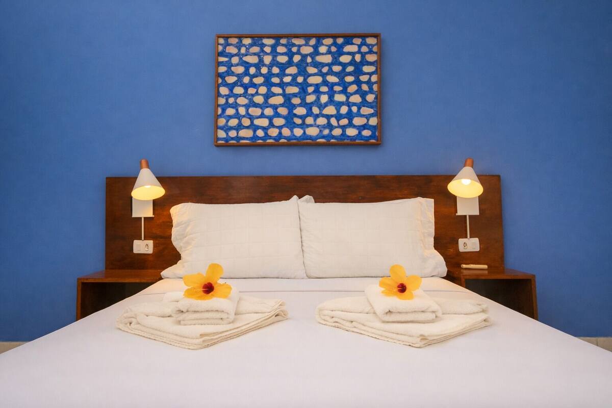 Cama bem arrumada com duas toalhas e uma flor, cabeceira de madeira, duas luminárias de mesa e parede azul com uma obra de arte estampada.