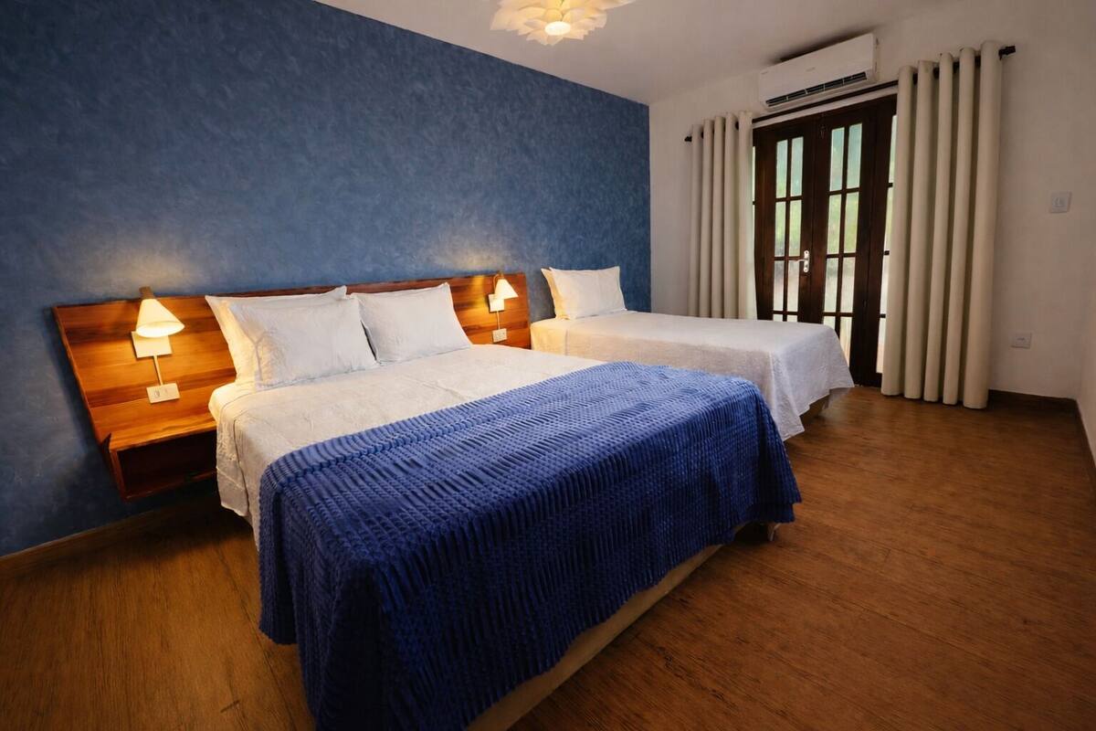Quarto de hotel com duas camas, cabeceiras de madeira, parede azul e uma janela com cortinas.