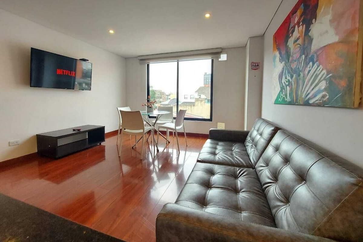 Uma sala de estar moderna com sofá de couro, televisão de tela plana fixada na parede, mesa de jantar com cadeiras e uma grande janela com vista para a cidade.