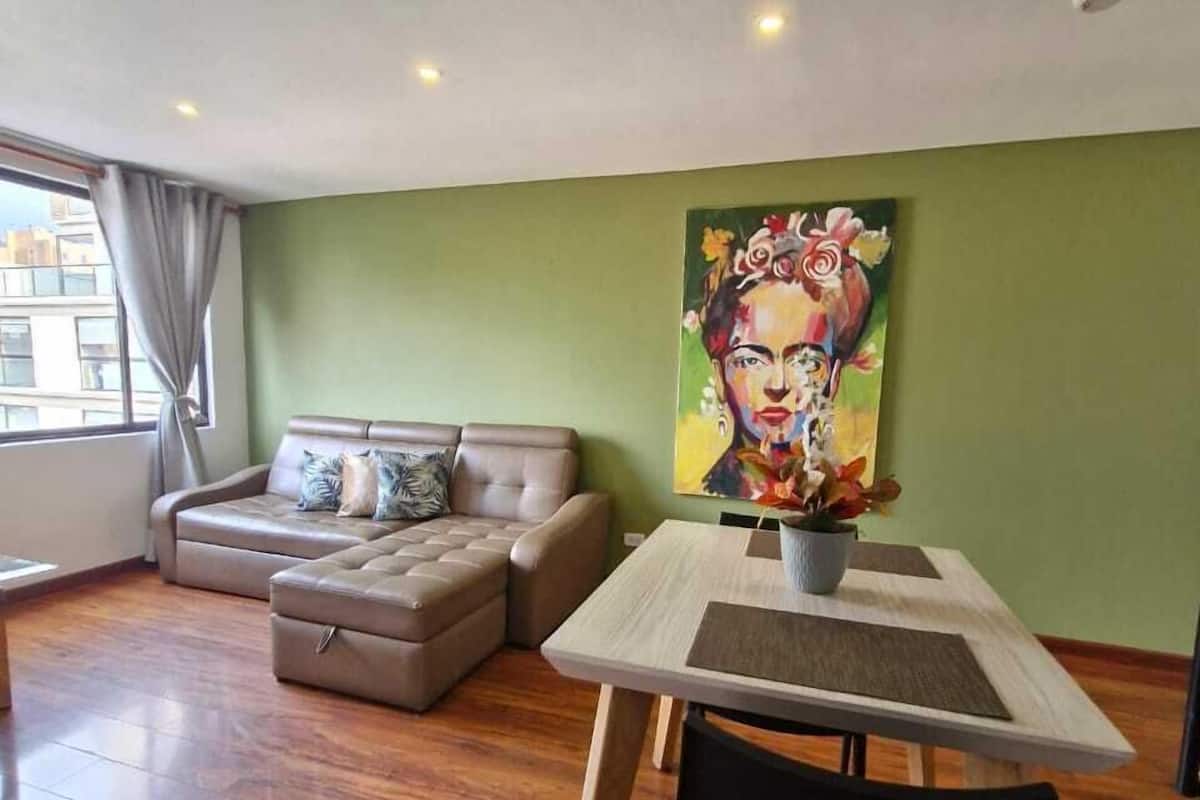 Uma sala de estar com um sofá, uma mesa de jantar com uma cadeira, um quadro na parede e uma janela com cortinas.