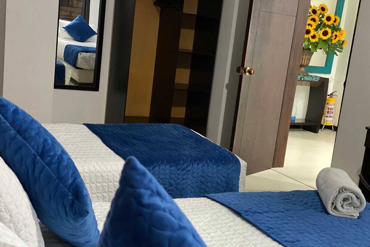 Quarto de hotel com duas camas, cada uma com roupa de cama azul e branca, um espelho e uma porta que dá para um corredor com extintor de incêndio.