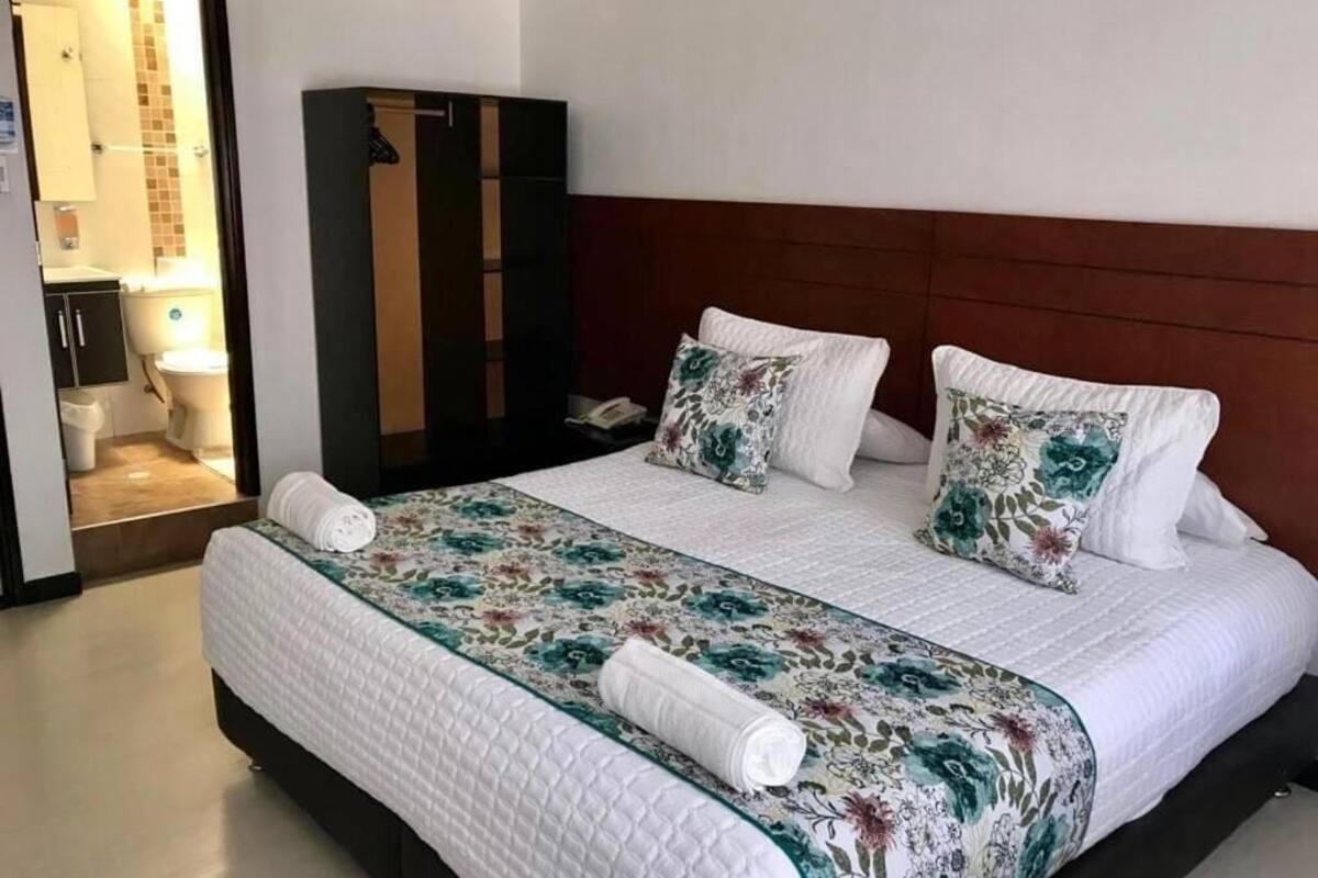 Cama bem arrumada com roupa de cama e almofadas florais, cabeceira de madeira e banheiro visível com vaso sanitário e pia.