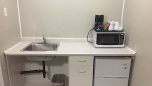 Uma kitchenette compacta com pia, micro-ondas, pequeno frigorífico e um cesto de lixo.