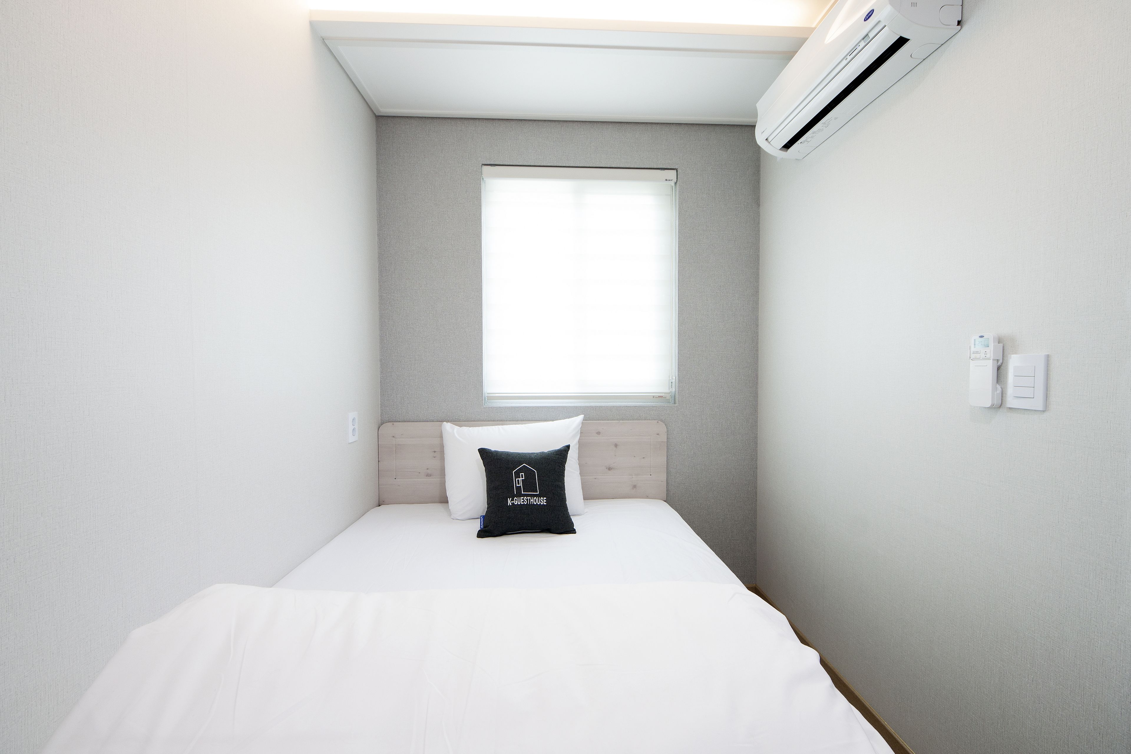 Quarto de hotel com cama, janela com persianas, unidade de ar condicionado e interruptor de luz fixado na parede.