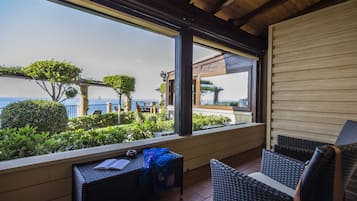 Uma área de estar coberta ao ar livre com móveis de vime, uma mesa com um livro e um casaco, e vista para o mar e vegetação.