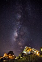 Céu estrelado à noite, com a Via Láctea visível, uma casa moderna com varanda, e uma árvore em primeiro plano.