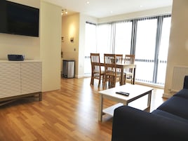Uma sala de estar com televisão de tela plana, uma mesa de jantar com cadeiras, um sofá e uma mesinha com um controle remoto.