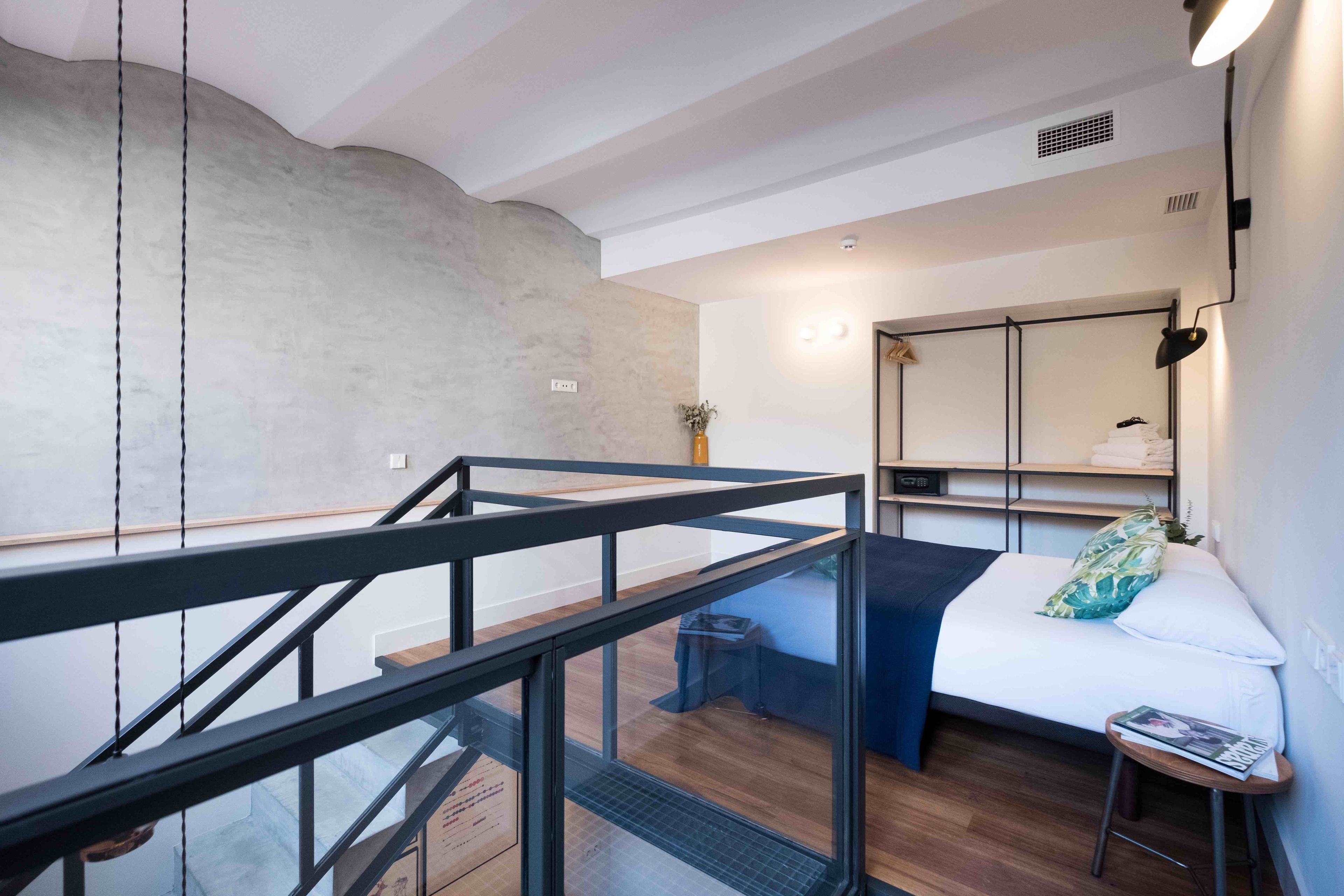 Quarto moderno com cama, mesa de cabeceira e um pequeno banquinho. Uma escada com corrimão de vidro leva a um andar superior.