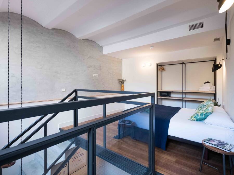 Quarto moderno com cama, mesa de cabeceira e um pequeno banquinho. Uma escada com corrimão de vidro leva a um andar superior.