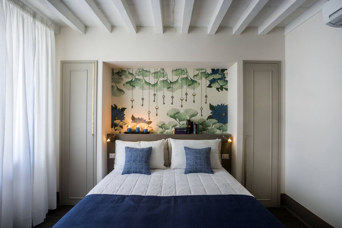 Quarto com cama, duas almofadas, cabeceira com enfeites pendurados e parede com padrão de folhagem.