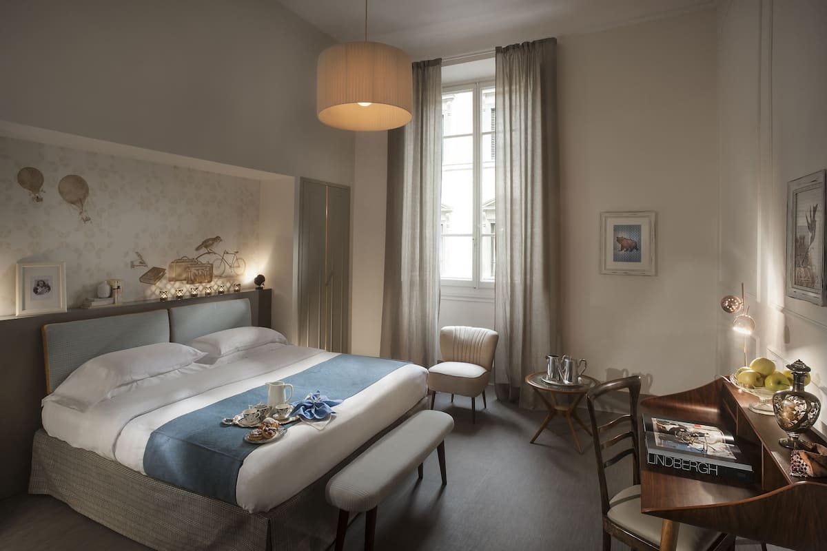 Quarto de hotel com uma cama grande, uma escrivaninha com uma revista, uma cadeira e uma janela com cortinas.