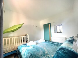 Quarto com berço, cama com colcha azul e branca, porta verde e janela com espelho.