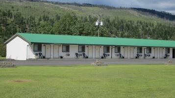 Um motel com teto verde e fachada branca, com uma fileira de quartos individuais, cada um com uma cadeira, e uma pequena área de jardim na frente.