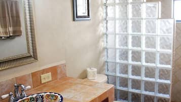 Casa de banho com lavatório revestido a azulejos, espelho, sanita e chuveiro com painéis de vidro jateado.