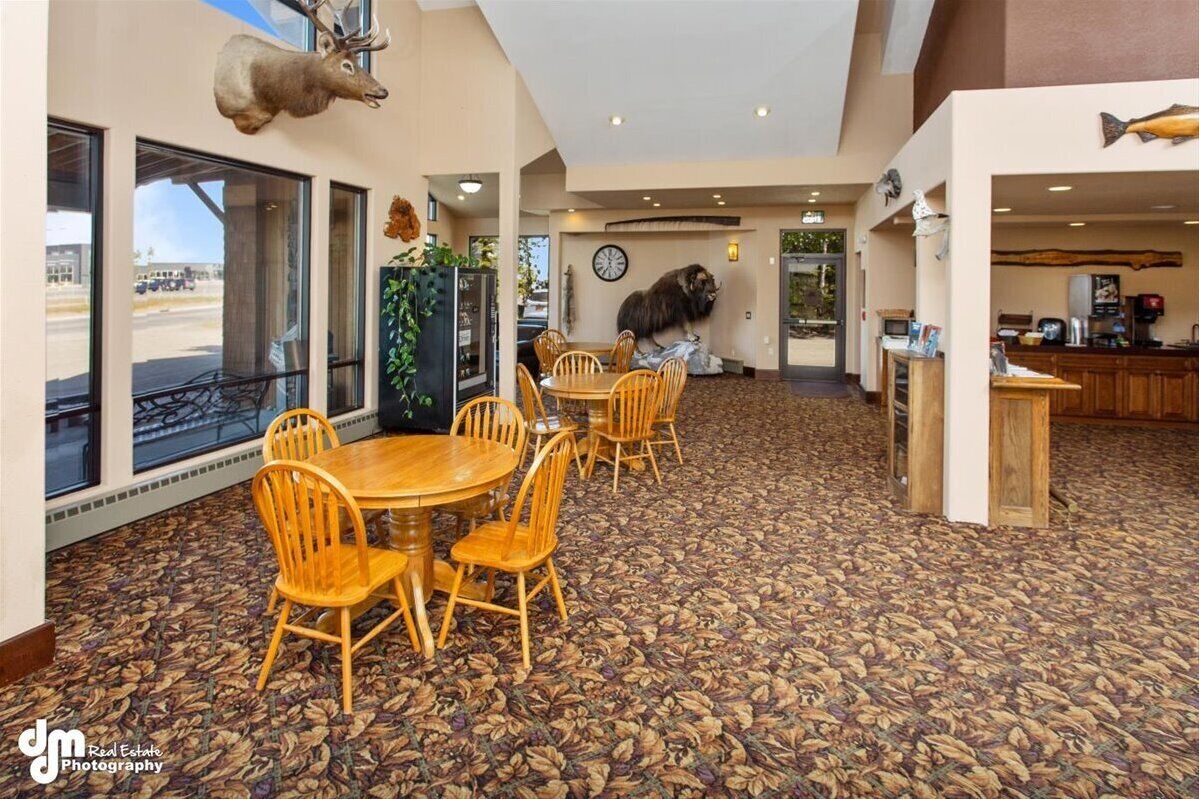 Uma sala de jantar com uma mesa de madeira redonda e cadeiras, um relógio na parede, e uma janela grande com vista para o exterior.