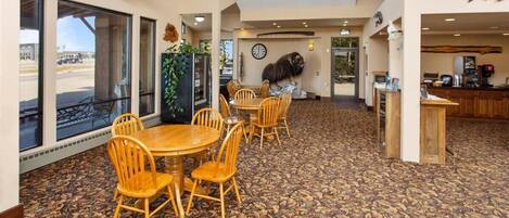Uma sala de jantar com uma mesa de madeira redonda e cadeiras, um relógio na parede, e uma janela grande com vista para o exterior.