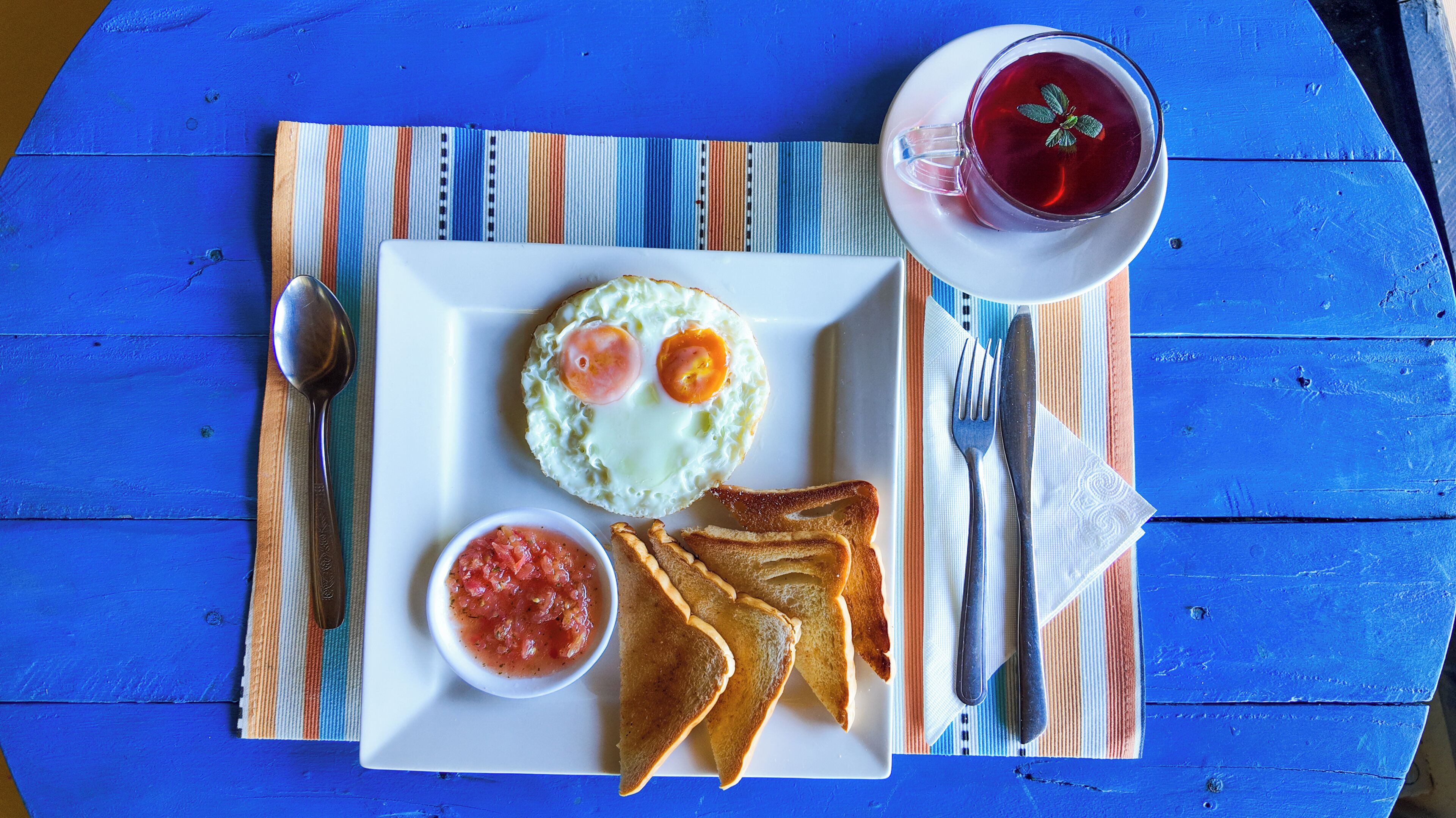 Prato de café da manhã com ovos, torradas e molho de tomate sobre uma mesa azul.