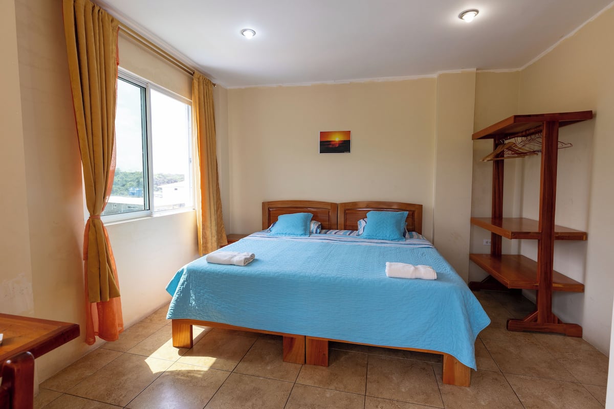 Quarto de hotel com uma cama grande, cabeceira de madeira, uma janela com cortinas, um pequeno quadro na parede e uma estante de madeira.