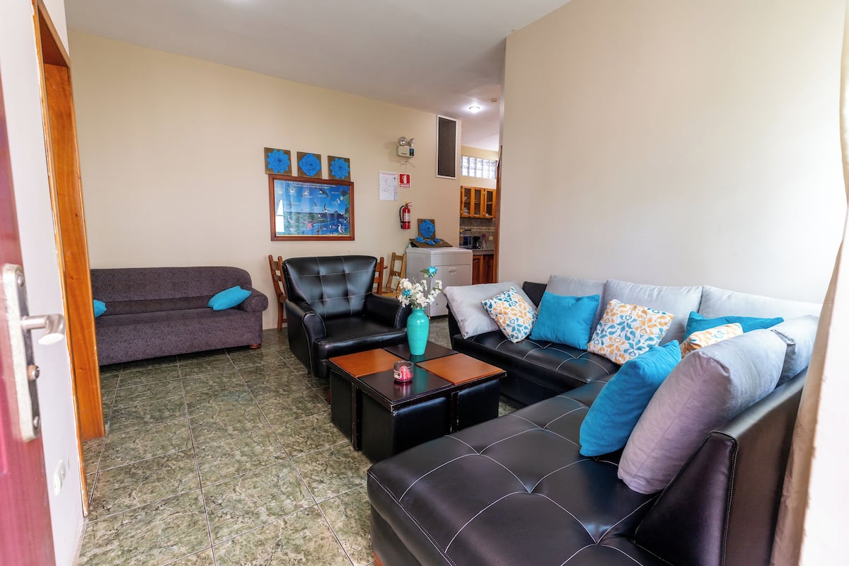 Sala de estar com sofá, poltrona e mesa de centro. Há um quadro na parede e um extintor de incêndio.