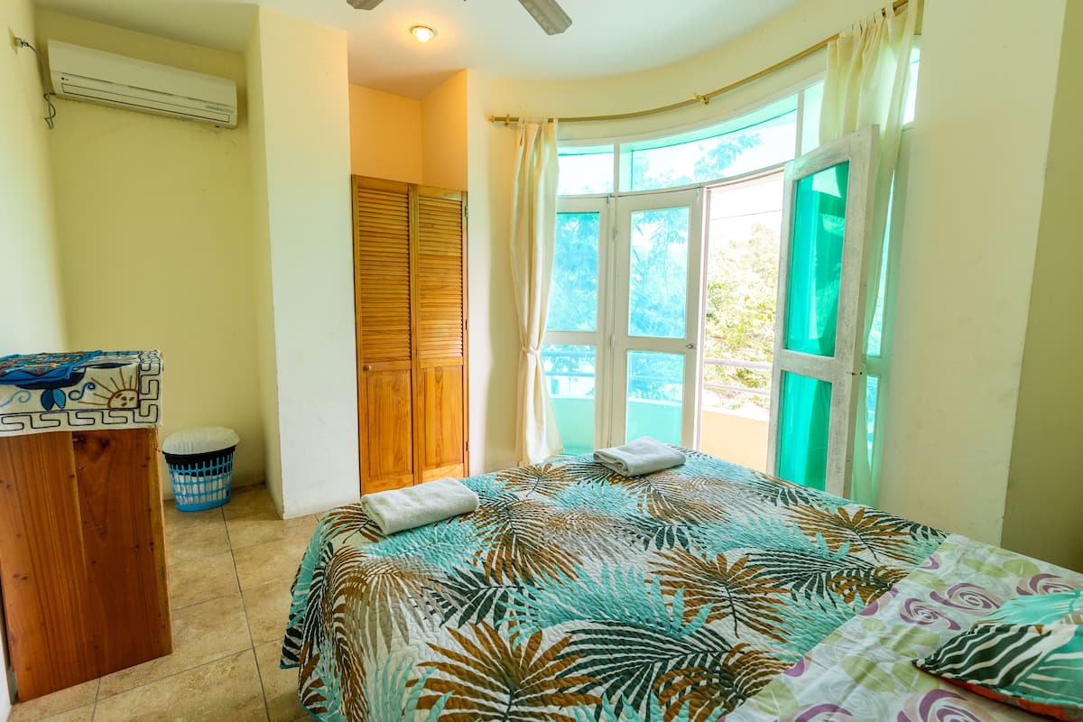 Quarto com cama, guarda-roupa de madeira, cesto de roupa azul e janela com cortinas.