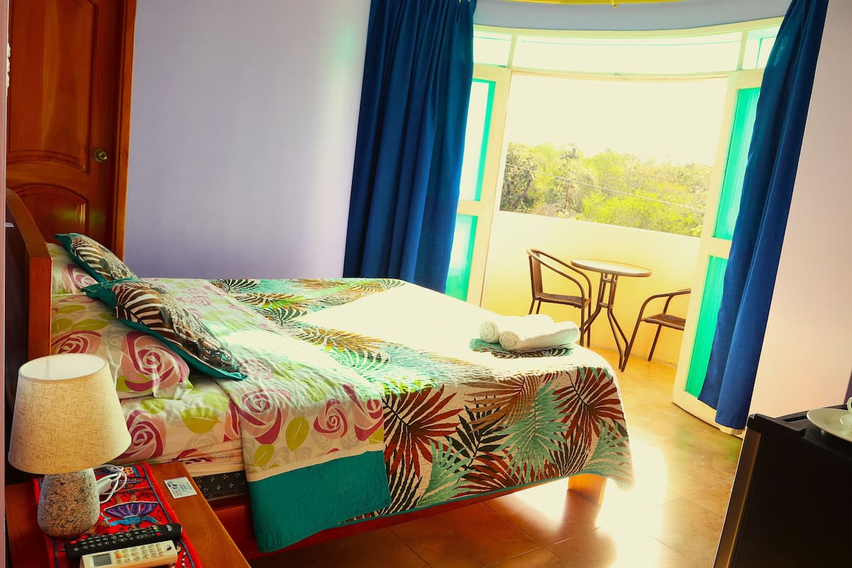 Quarto de hotel com cama, escrivaninha com luminária, cadeira e vista para o exterior.