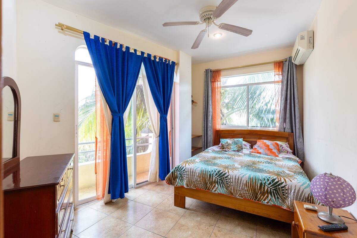 Quarto com cama, ventoinha no teto, janela com cortinas, penteadeira e mesa de cabeceira com abajur.