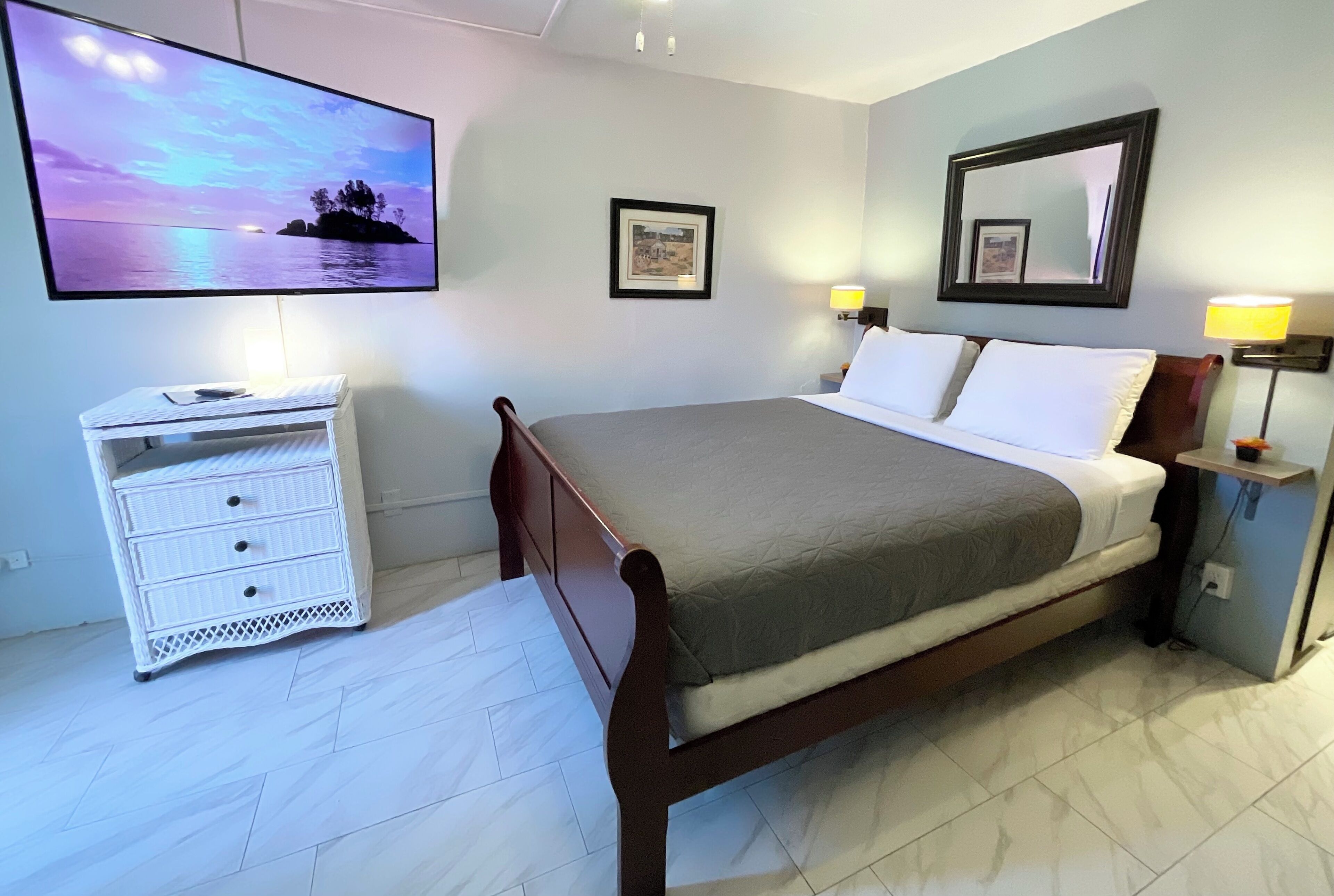 Quarto com cama, televisão na parede, cômoda e mesa de cabeceira com abajur.