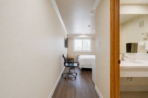 Quarto de hotel com cama, escrivaninha com cadeira, janela com persianas e banheiro com pia e espelho.