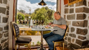 Um homem sentado à mesa com vista para um pátio e um edifício ao fundo.