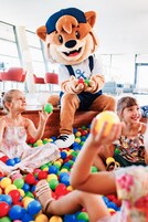 Um mascote com fantasia de cachorro sentado em uma superfície de madeira, cercado por crianças brincando com bolas coloridas em uma piscina de bolinhas.