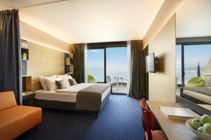Quarto de hotel moderno com uma cama grande, uma escrivaninha, uma cadeira, uma mesinha, um sofá, uma TV e vista para o oceano.