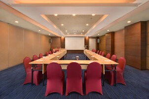 Uma sala de conferências com uma mesa comprida, cadeiras vermelhas e uma tela de projetor na frente.
