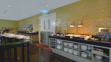 Uma área de buffet com mesa de vidro, máquina de café e diversas opções de comida.