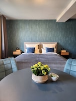 Quarto de hotel com uma cama grande, duas mesas de cabeceira, uma mesa redonda com um vaso de flores e uma cadeira.