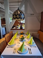 Uma sala de jantar com uma mesa posta para quatro pessoas, com guardanapos amarelos, um vaso de flores e uma luminária pendente geométrica.