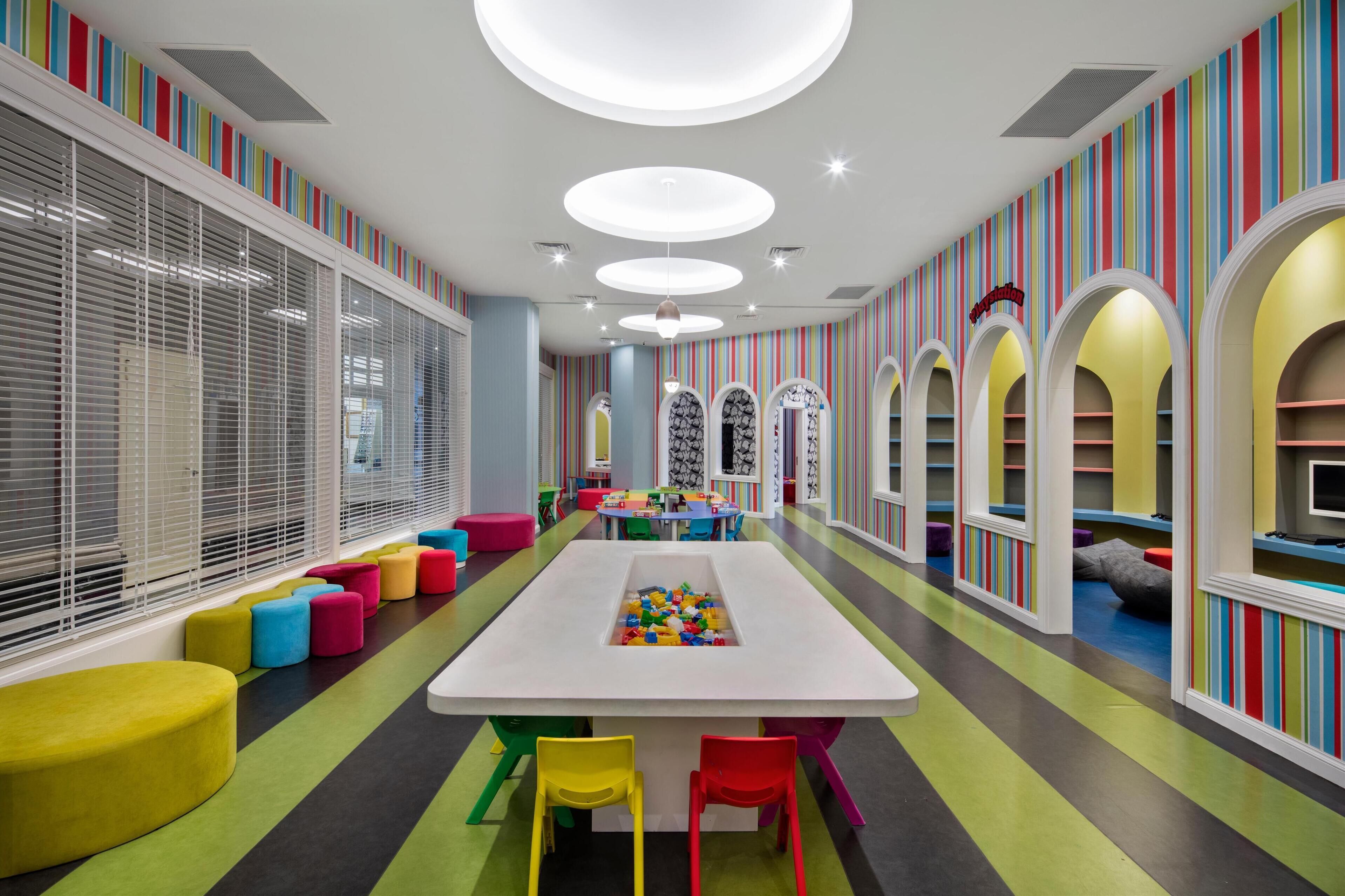 Uma área de recreação infantil vibrante, com uma mesa comprida, cadeiras coloridas e armários de armazenamento ao longo das paredes.