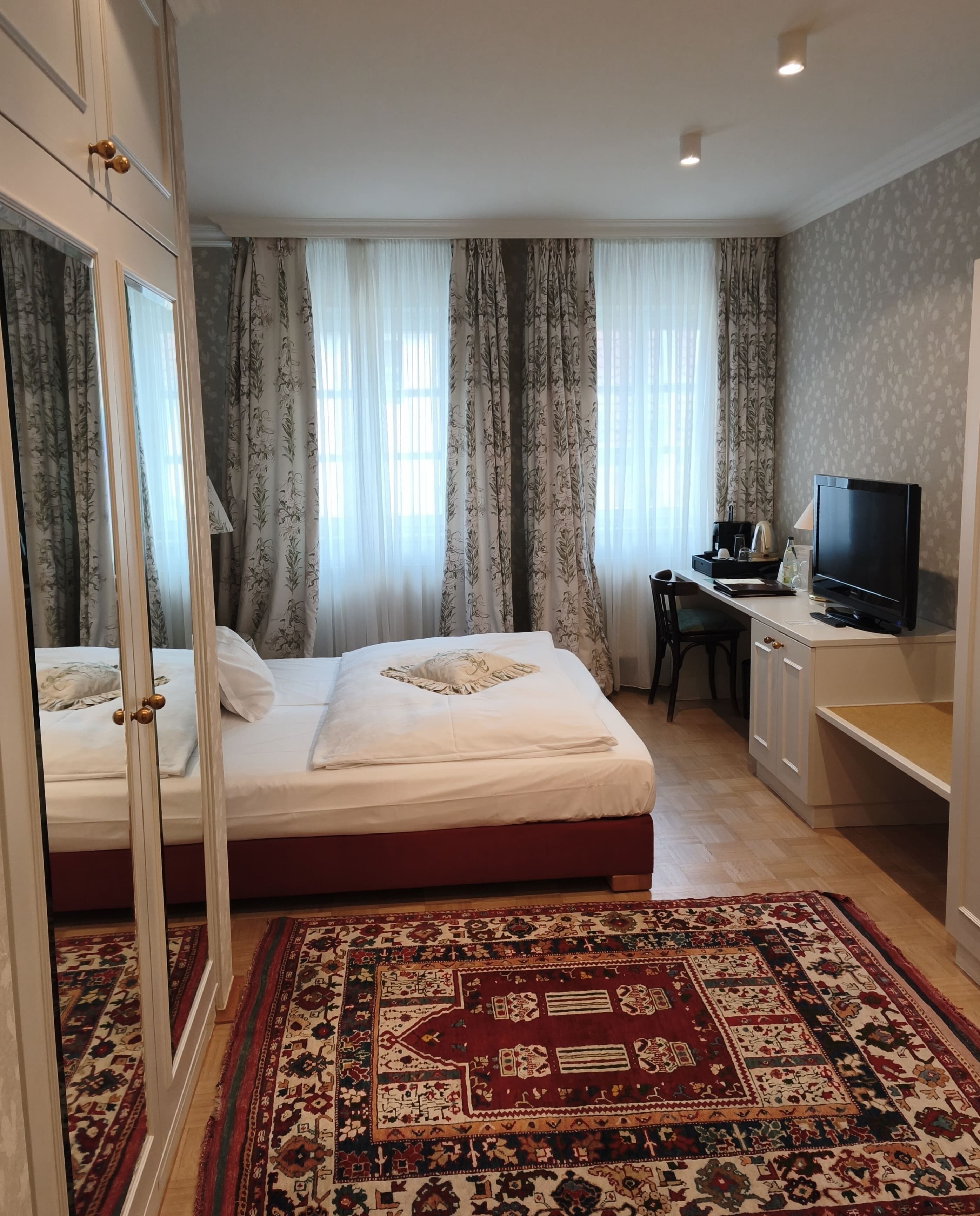 Quarto de hotel com cama, escrivaninha com televisão e um espelho grande.