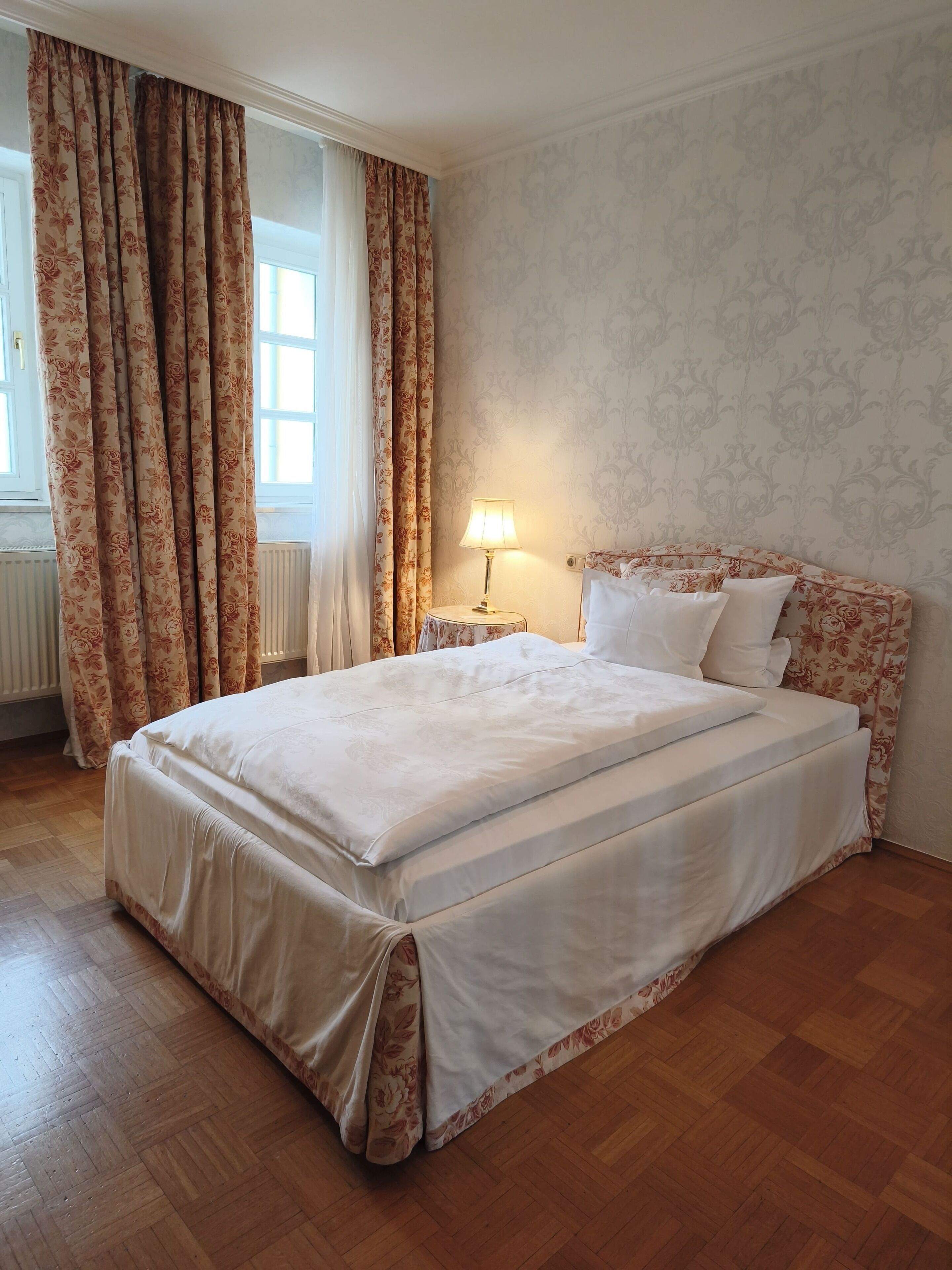 Uma cama bem arrumada com roupa de cama estampada, um abajur e uma janela com cortinas.