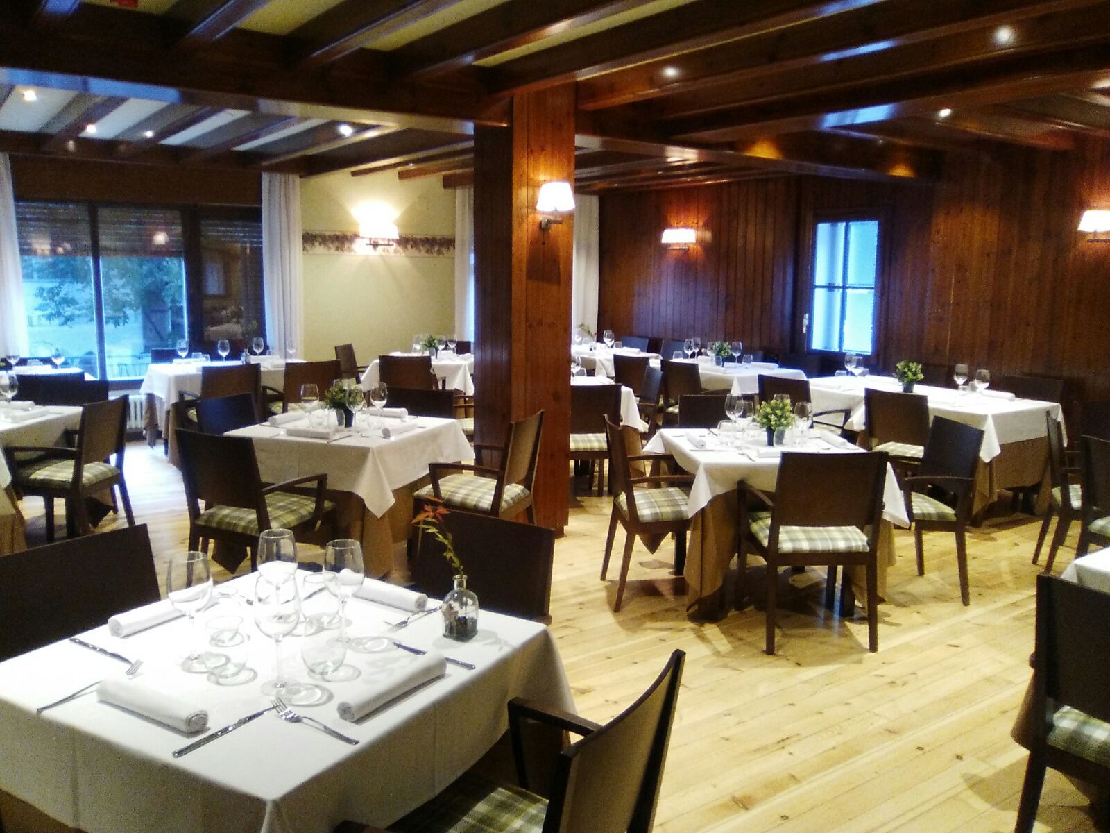 Um restaurante com várias mesas preparadas para refeições, piso de madeira e grandes janelas.
