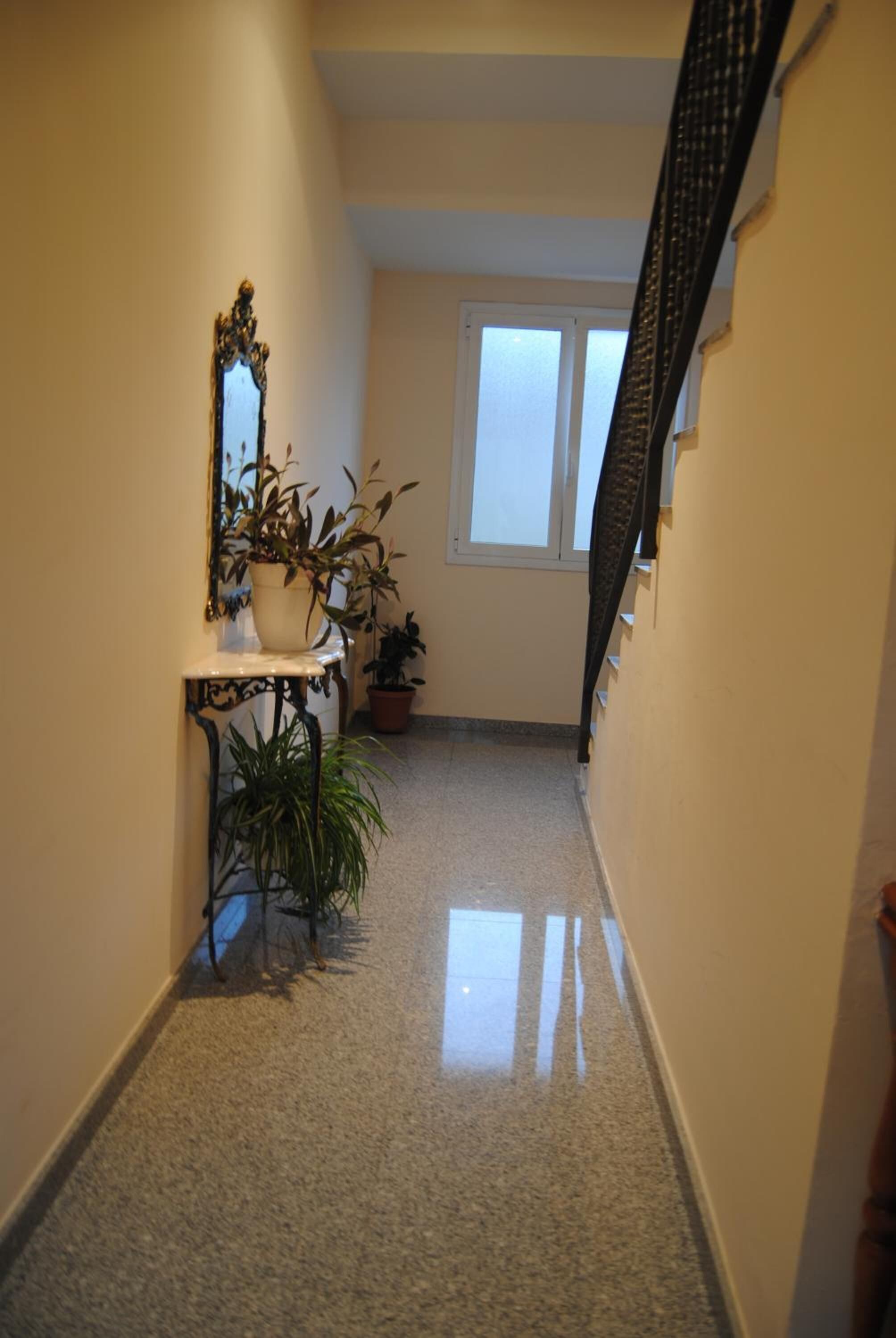 Um corredor estreito com uma escada à direita, uma janela no final e uma mesa com plantas e um espelho à esquerda.