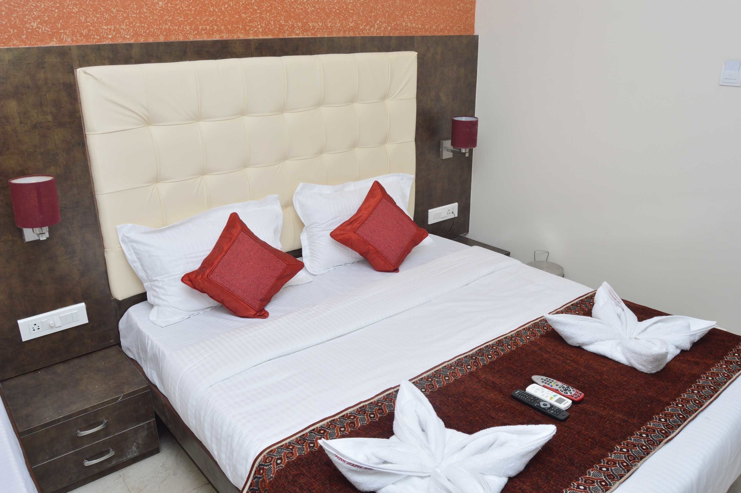 Quarto de hotel com uma cama grande, dois travesseiros vermelhos, um cobertor branco e uma toalha na cama. Há duas mesas de cabeceira com abajures e uma tomada.