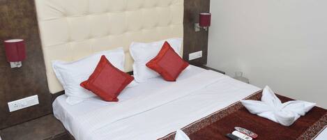 Quarto de hotel com uma cama grande, dois travesseiros vermelhos, um cobertor branco e uma toalha na cama. Há duas mesas de cabeceira com abajures e uma tomada.