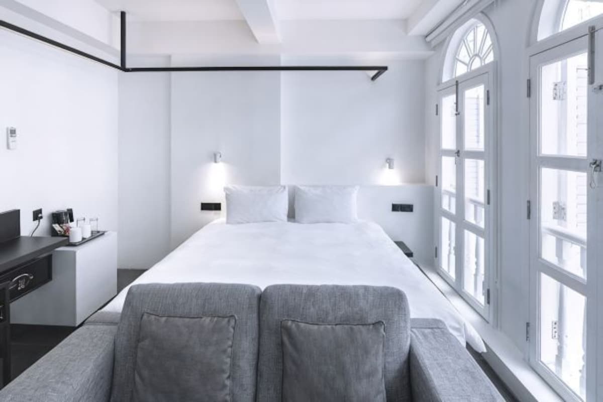 Quarto de hotel moderno com uma cama grande, abajures, uma escrivaninha com cadeira e janelas amplas.