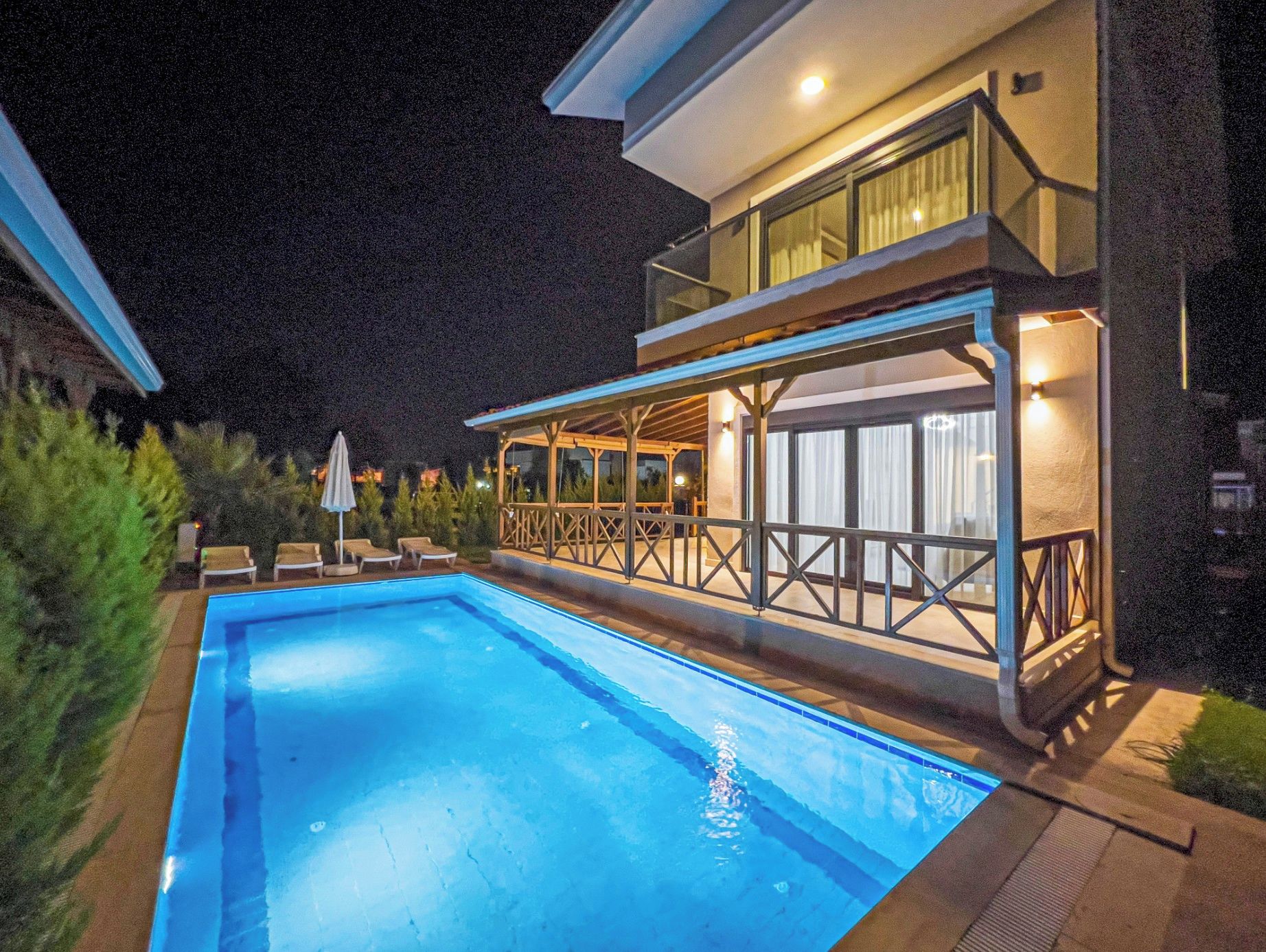 Piscina com área coberta, cercada por vegetação e céu noturno.
