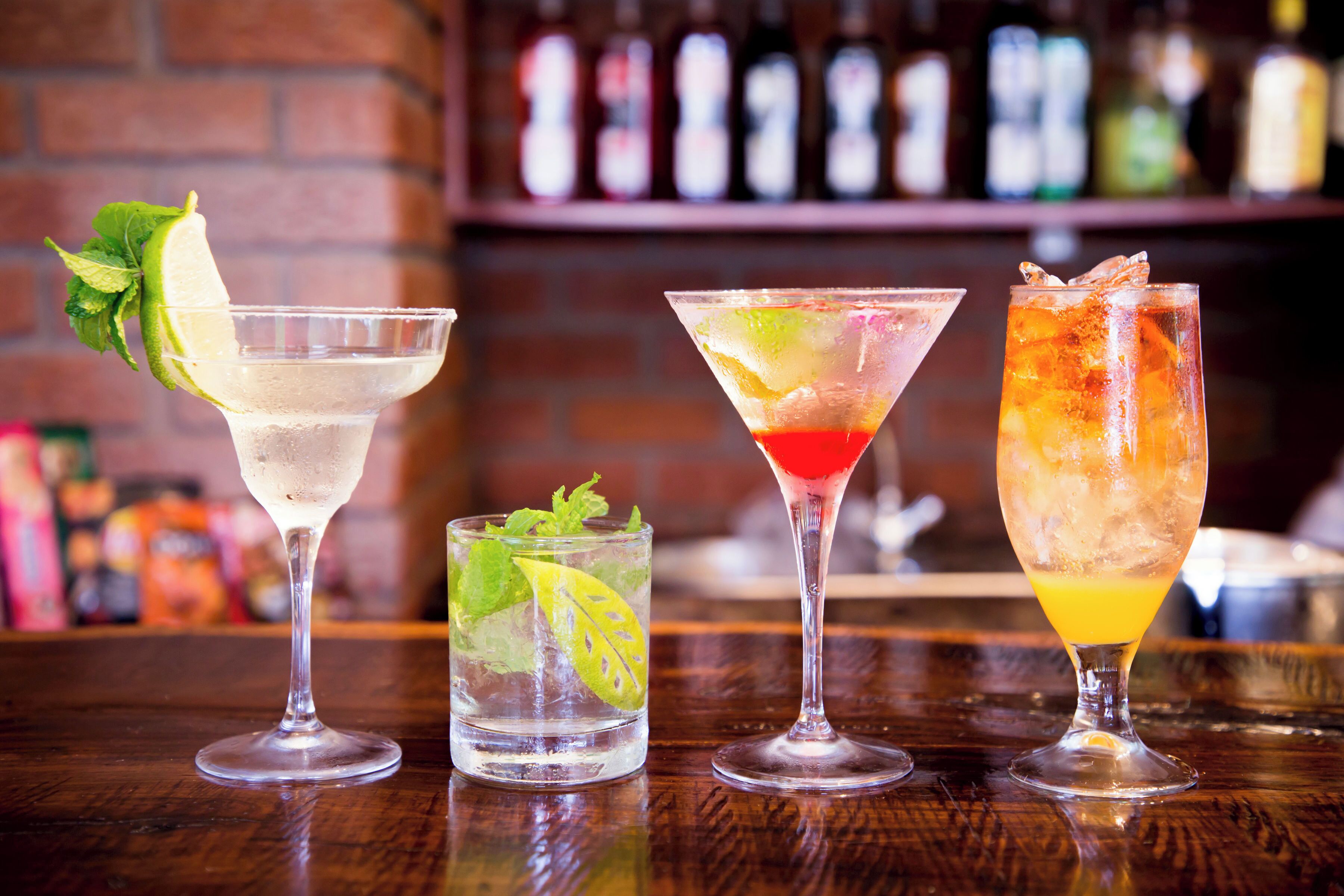 Três cocktails diferentes em um balcão de bar, com parede de tijolos ao fundo.