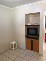 Quarto com televisão sobre suporte de madeira, um banquinho branco e um armário com uma mesa vermelha ao fundo.