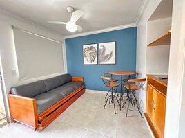 Um quarto com sofá cinzento, ventoinha no teto, duas obras de arte com folhas emolduradas, uma mesa redonda pequena com cadeiras e um armário de madeira.