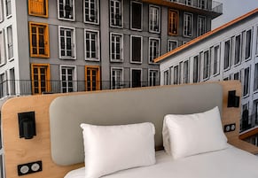 Quarto de hotel com cama, cabeceira de madeira e vista para um edifício com várias janelas e sacadas.