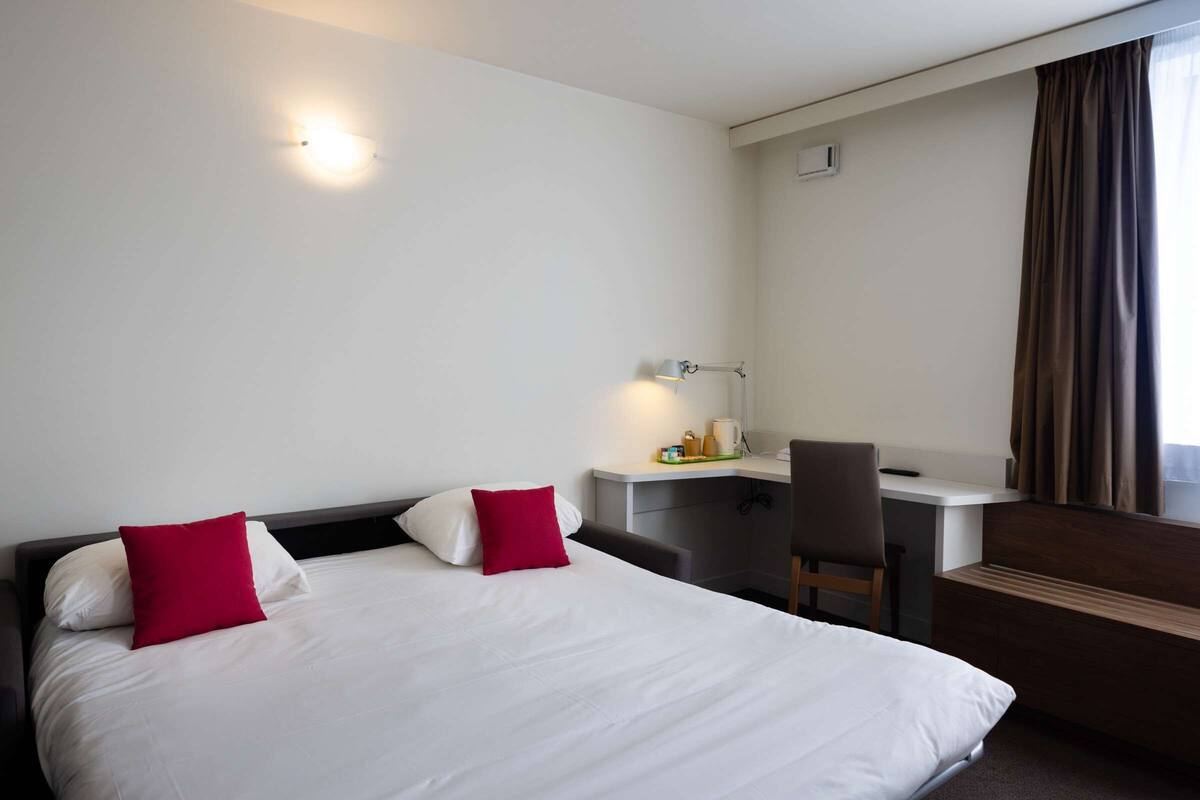 Quarto de hotel com cama, escrivaninha, cadeira e janela com cortinas.